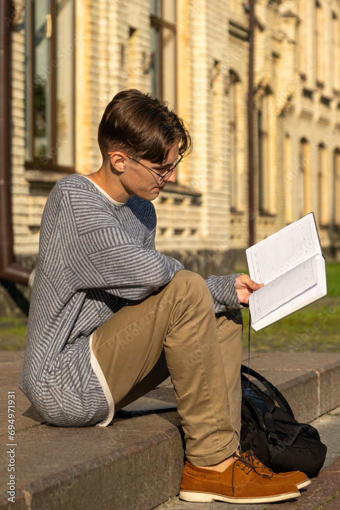 Young man with glasses sits on step reading notes in planner before ...