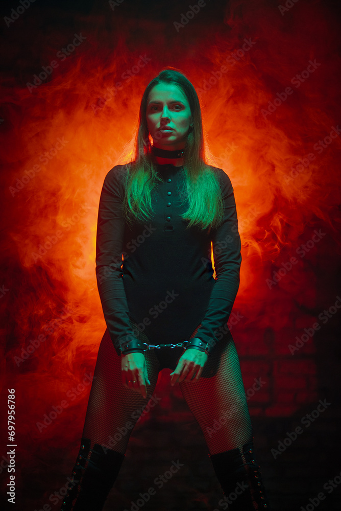 Beautiful girl in bodysuit with hands in handcuffs posing on the red light smoke background ...