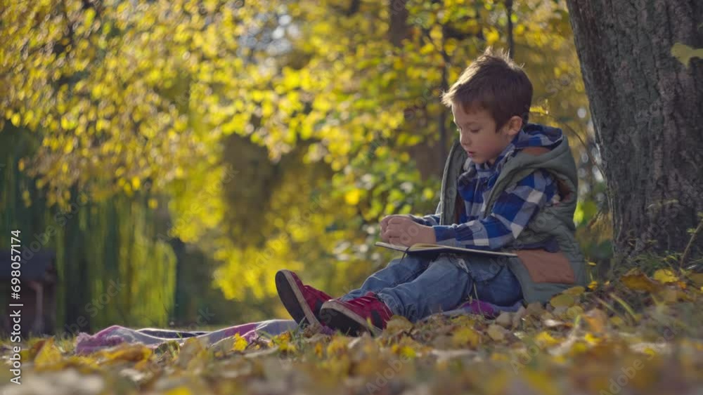 A boy writes homework in a notebook sitting on a yellow leaf in autumn ...