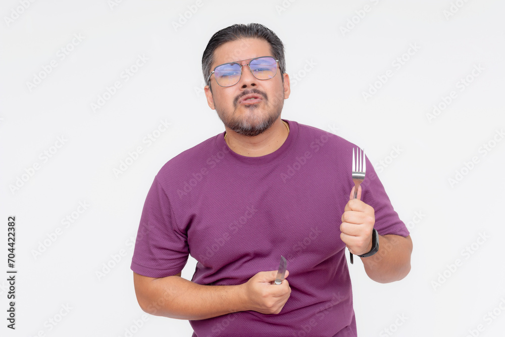 A hungry middle aged man holding a fork and knife feeling his stomach ...