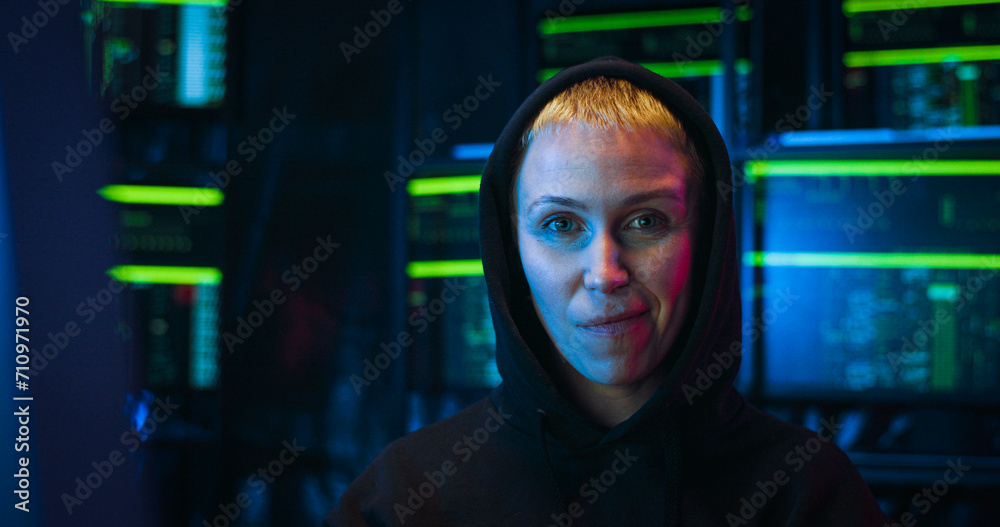 Caucasian female hacker in dark hood looking to camera on spinning chair and looking straight at ...