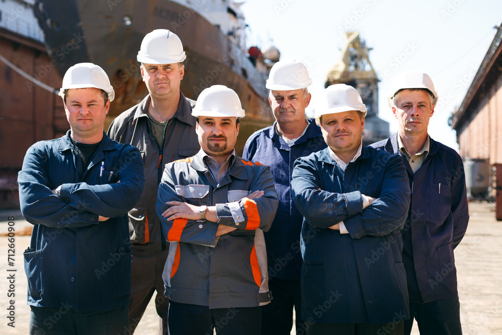 Shipyard workers at float dock wearing hard hats, stand arms-crossed ...