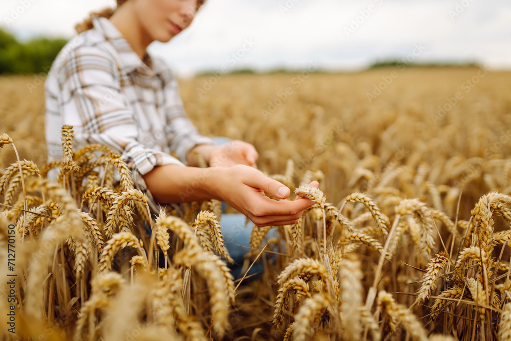 Young woman farmer walking across field and running her hand through golden ears of wheat ...