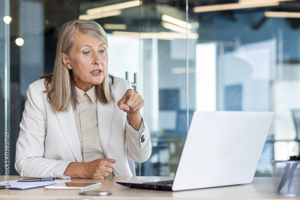 Angry senior business woman sitting in the office at the desk in a suit ...