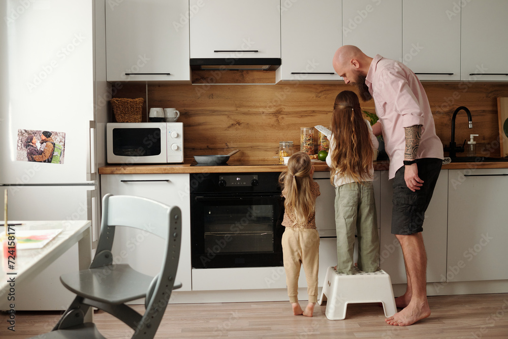 Two little siblings standing by kitchen counter next to their father ...