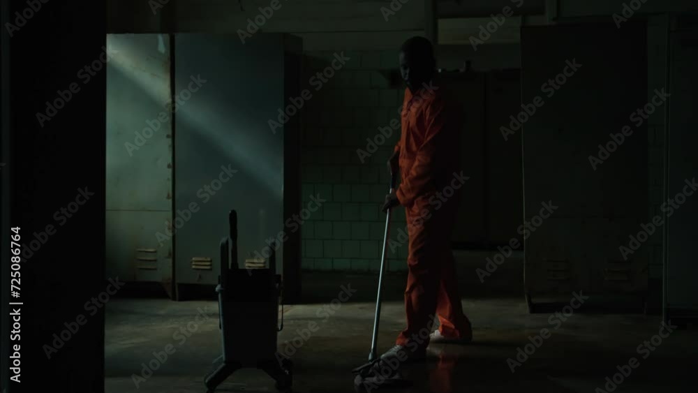 Male inmate in prison uniform cleaning floor in dark room with mop ...