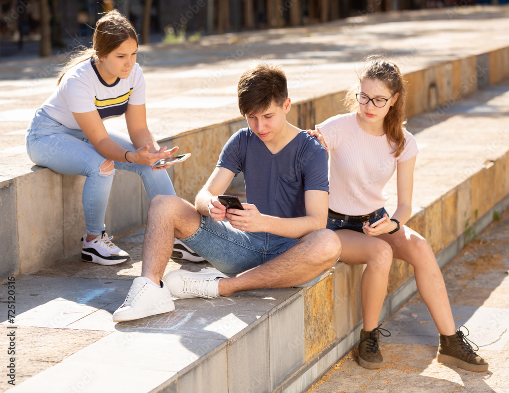 Interested teen boy sitting with phone on summer city street with two ...