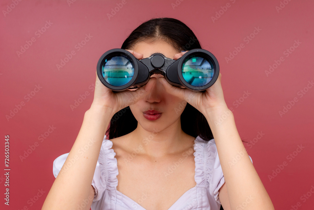 Portrait of a focused Asian woman gazing through binoculars against a ...