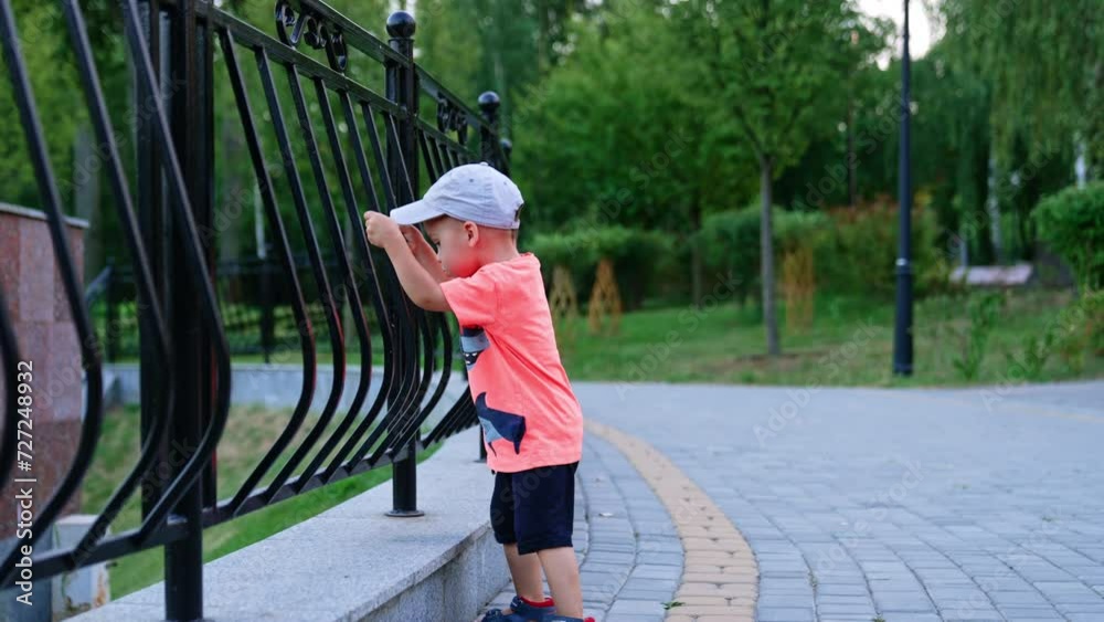 Toddler stands on the edging holding by the fence. Curious kid looks ...