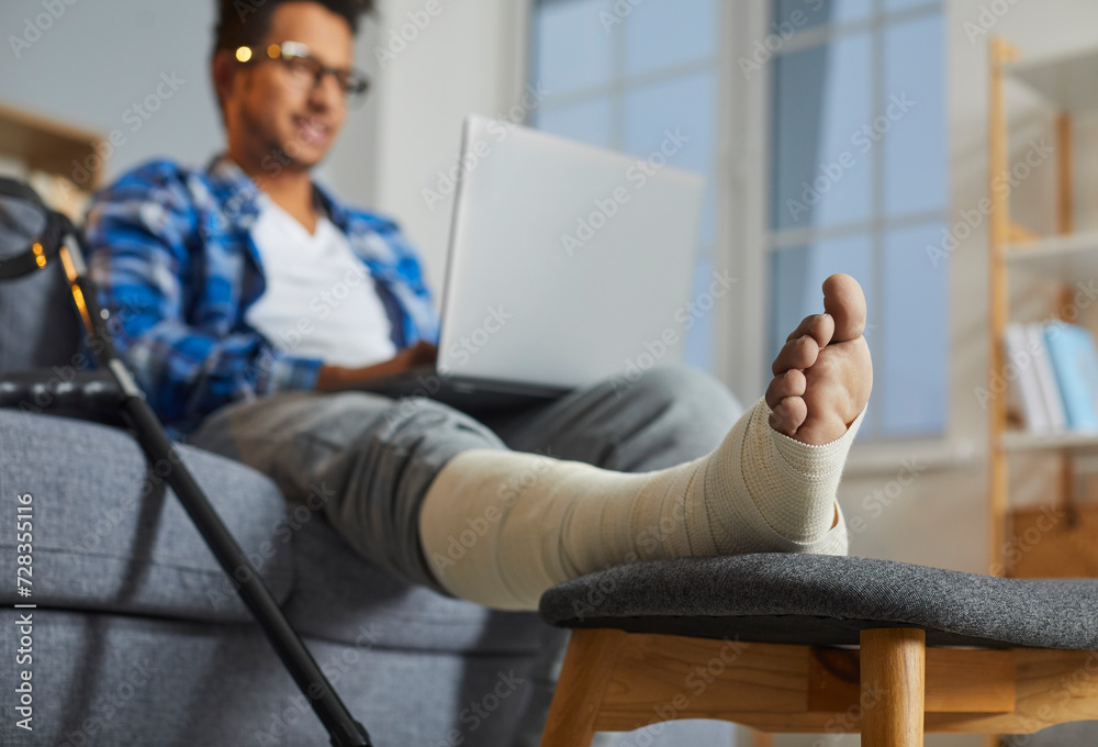 Young man with broken leg sitting on sofa using laptop. Man with ...