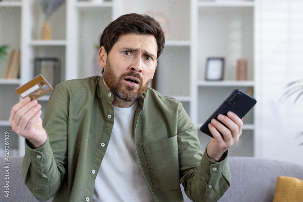 Confused adult man holding smartphone and bank card in hands while ...