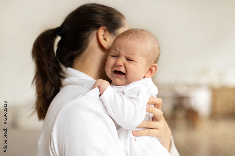 Young mother holding crying newborn baby girl or boy, calming kid, lullying in bedroom interior ...