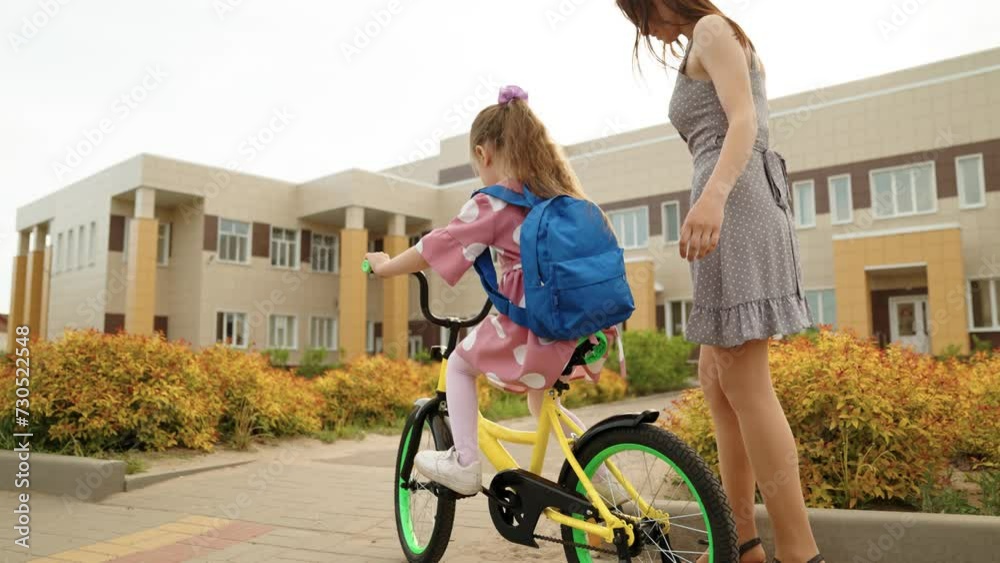 Playful little girl pupil riding on bike at schoolyard with mother ...