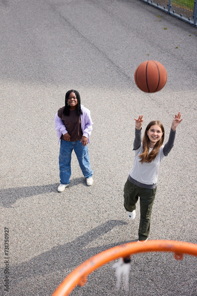 Teenage girl throwing ball in basket by female friend at sports field ...