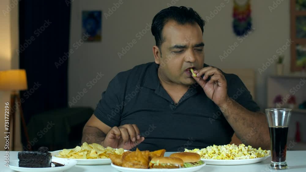 A middle-aged unhealthy hungry man enjoying a variety of snacks at home ...