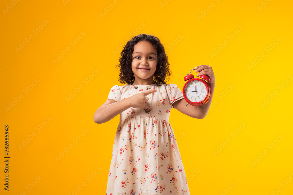 Kid girl holding an alarm clock in hand and pointing finger at it. The ...