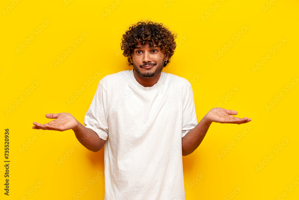 unsure curly indian man shrugs his shoulders and raises his hands over ...