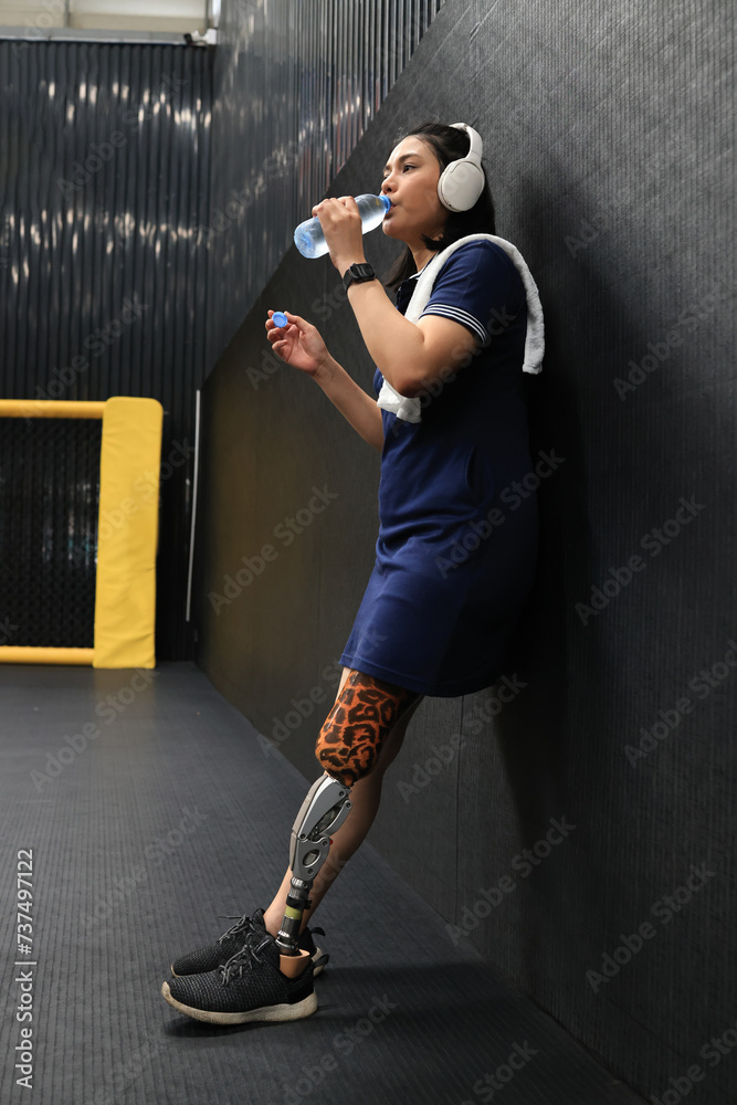 Sportswoman with prosthetic leg standing lean the wall and drinking ...