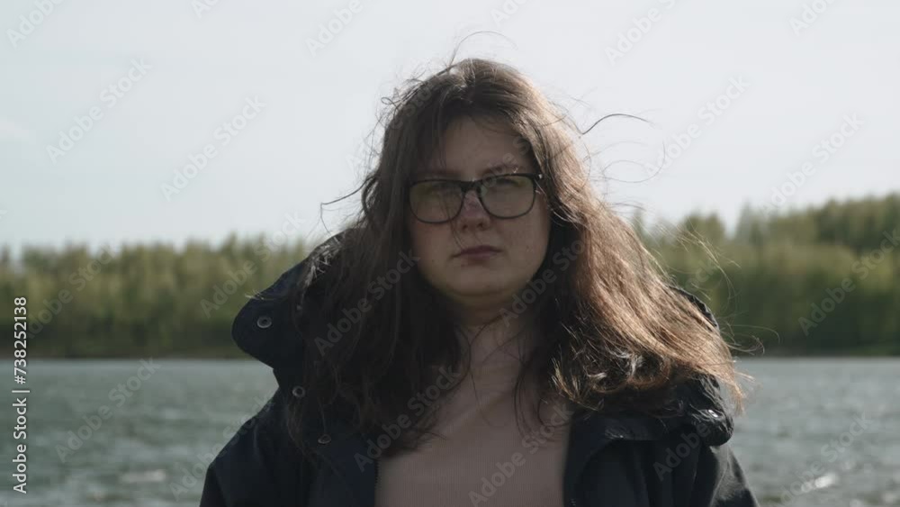 Portrait of sad woman in jacket stands in cold wind on river bank. Wind ...