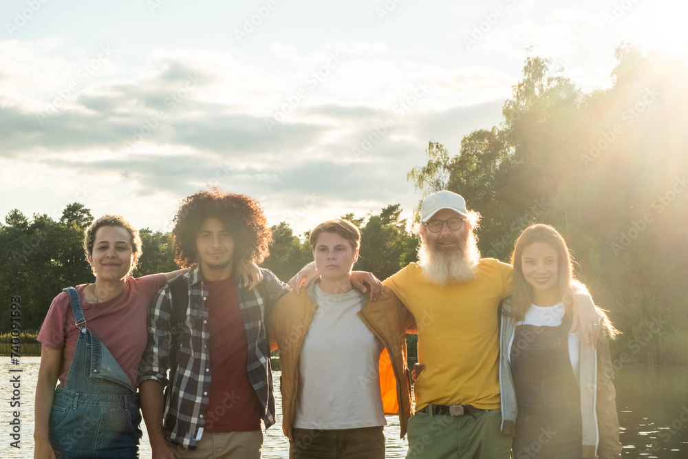 A group of friends stands together by a lake, backlit by the soft, warm ...