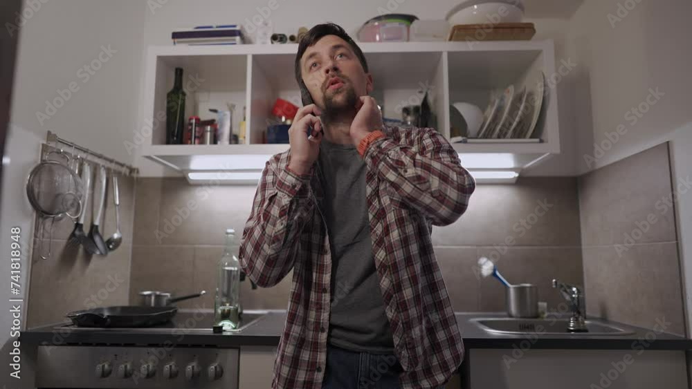 Sad man talking on a mobile phone at home in the kitchen. A puzzled ...