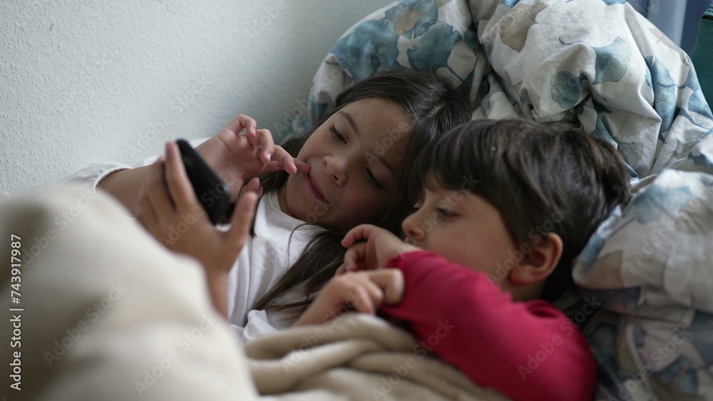 Children looking at cellphone device lying in bed under bedsheets ...