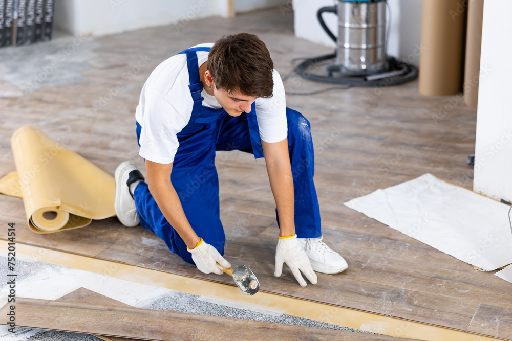 Skilled young carpenter using rubber mallet to lay click-lock laminate ...