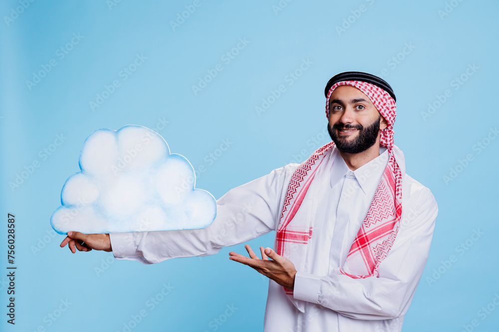 Smiling muslim man holding cloud shape and pointing with hand studio ...