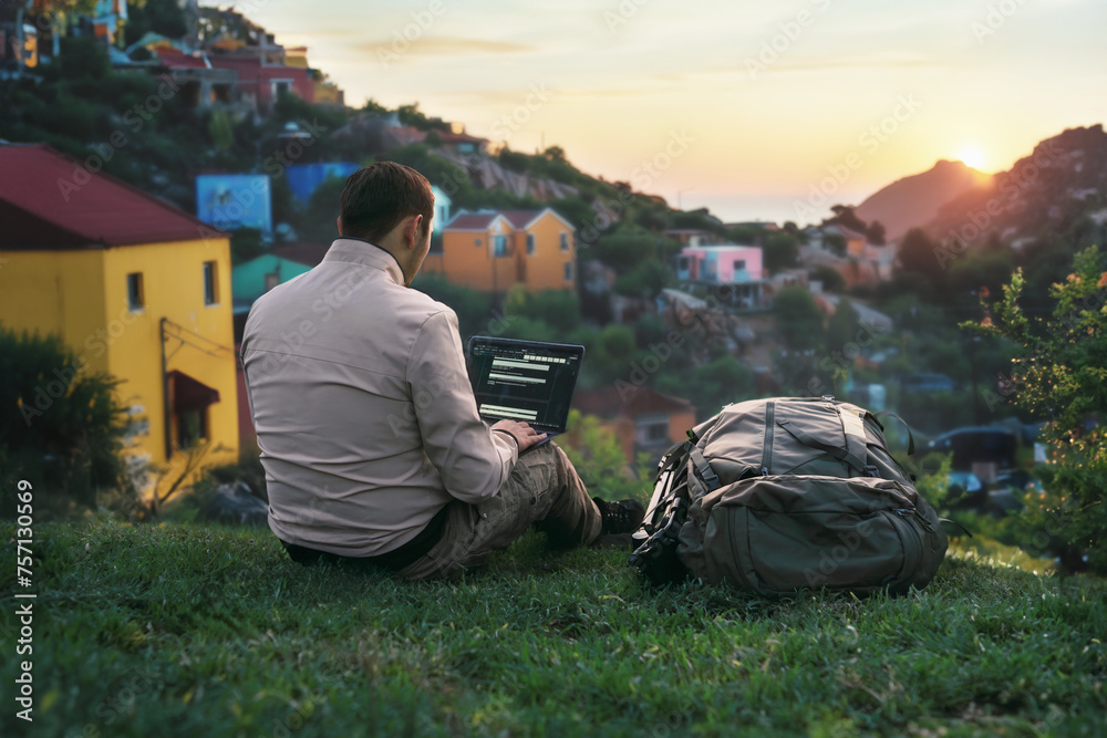 A digital nomad sits on a grassy hill overlooking a scenic village at ...