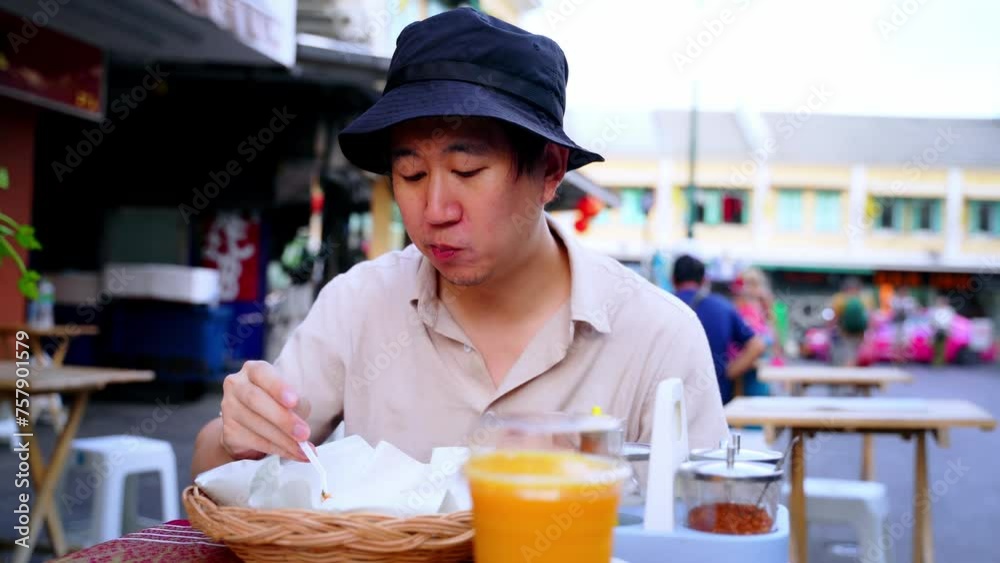 Young adult Asian man enjoys eating a local Thai food in the street food stall shop in Bangkok ...