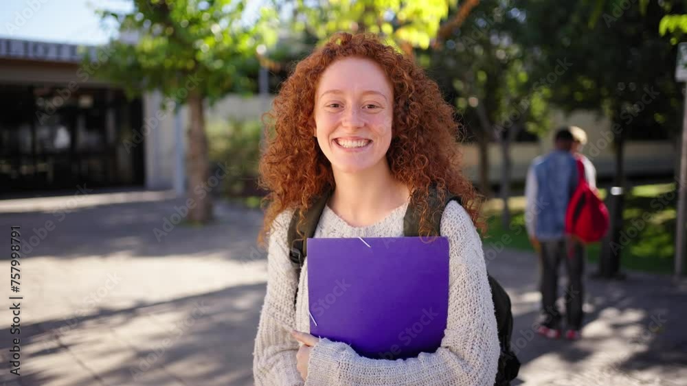 A young red-haired student smiles looking at the camera holding a ...