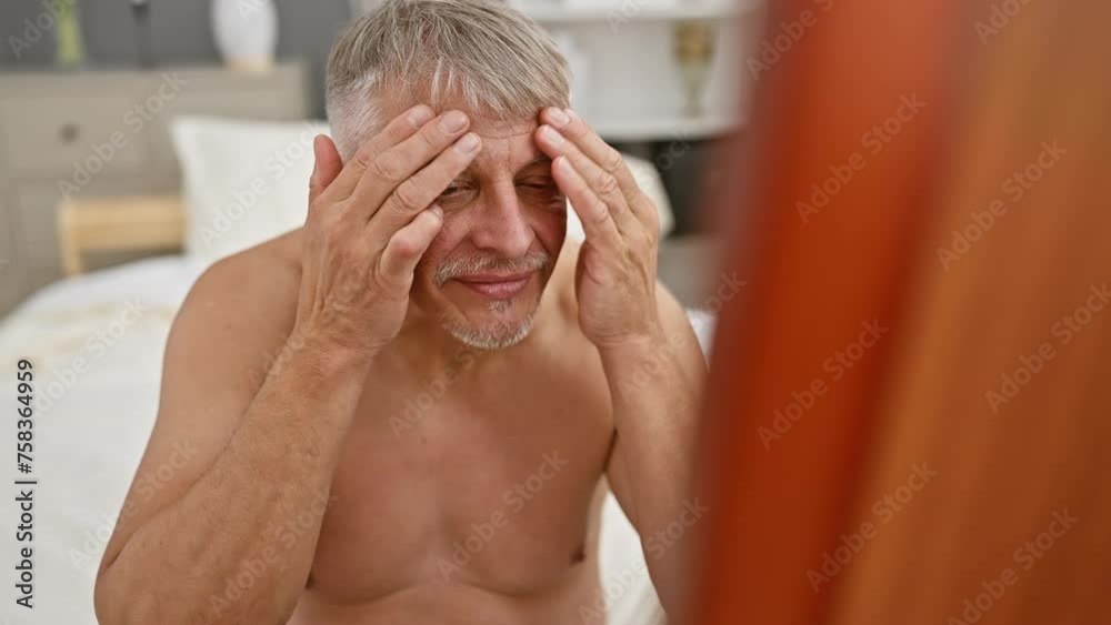 A shirtless, mature man examines his face in the bedroom mirror ...