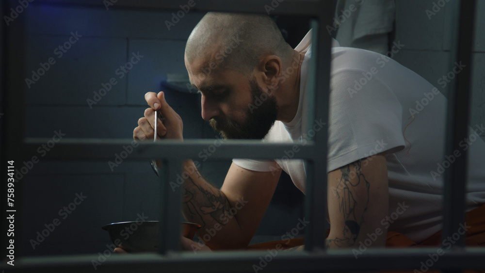 Inmate in tattoos in orange uniform sits on bed in jail cell, eats ...