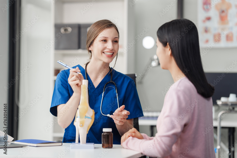 Caucasian female doctor explain to asian female patient using knee bone model at desk in medical ...