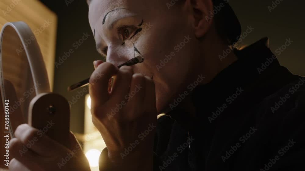 Chest up of mature female mime artist looking in mirror and painting ...