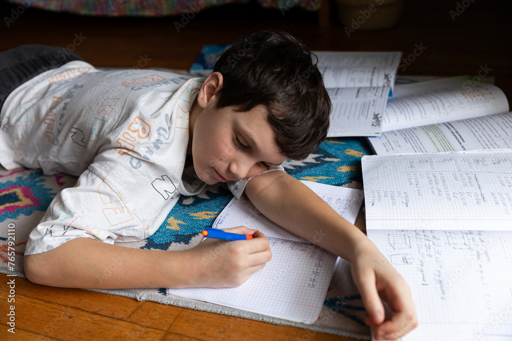 Teenage boy doing homework in his room lying on the floor, he writes in ...