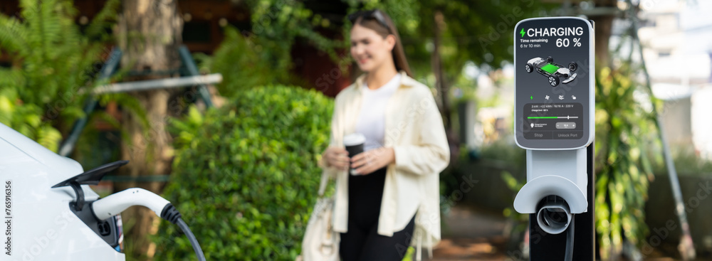 Young woman with coffee cup and sustainable urban commute with EV electric car recharging at ...