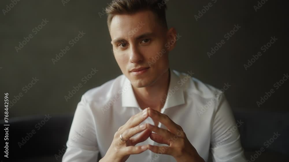 Confident young man in a white shirt, sitting with hands clasped ...