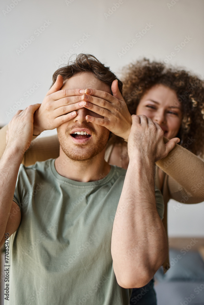 Adoring couple, curly young woman covering eyes of brunette man ...