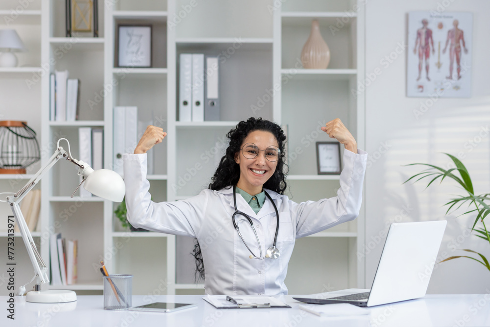 A joyful latino woman doctor with curly hair raises her arms in a ...