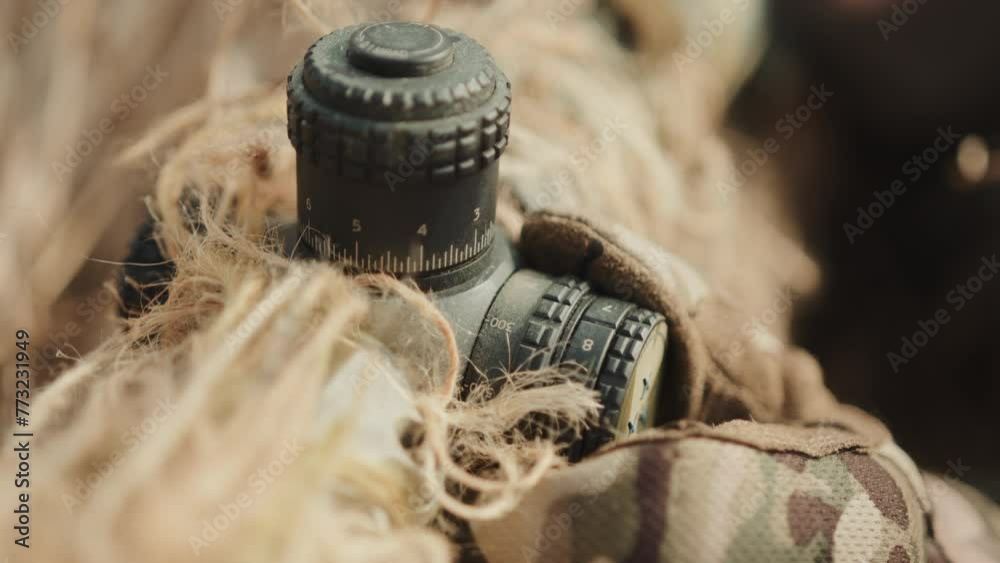 A gloved hand delicately adjusts the optic sight of a sniper rifle, a ...