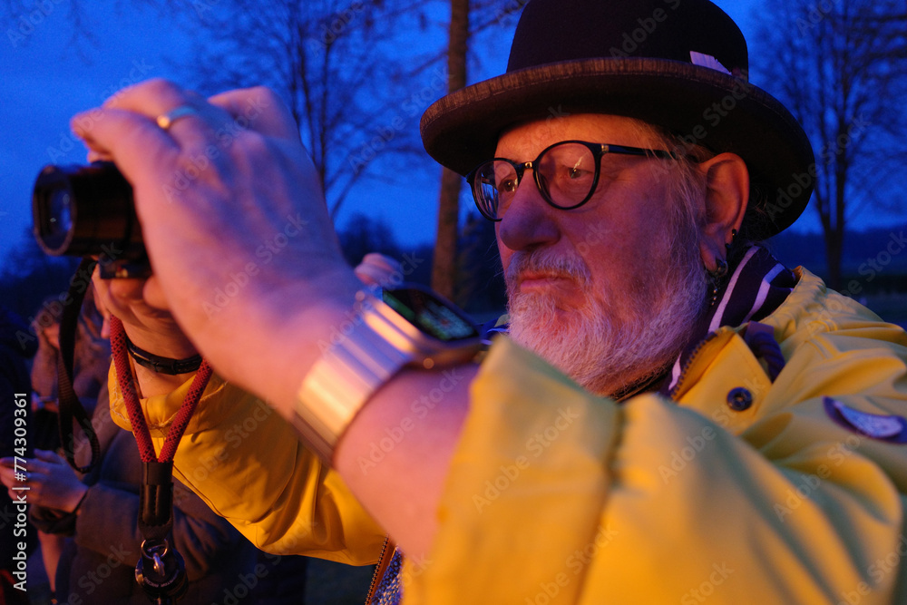 Older male vlogger in hat films the Easter bonfire. His face is lit red ...