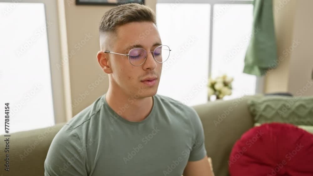 A contemplative young man with glasses sits indoors, giving a sense of ...
