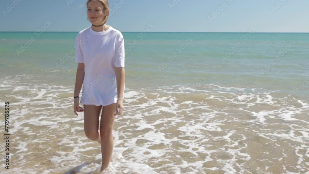 Pretty smiling teenage girl in a T-shirt walks at ocean shore, sea surf ...