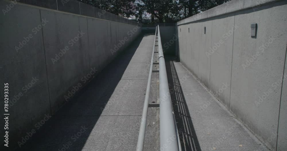 The image features a narrow, concrete pedestrian ramp flanked by high ...