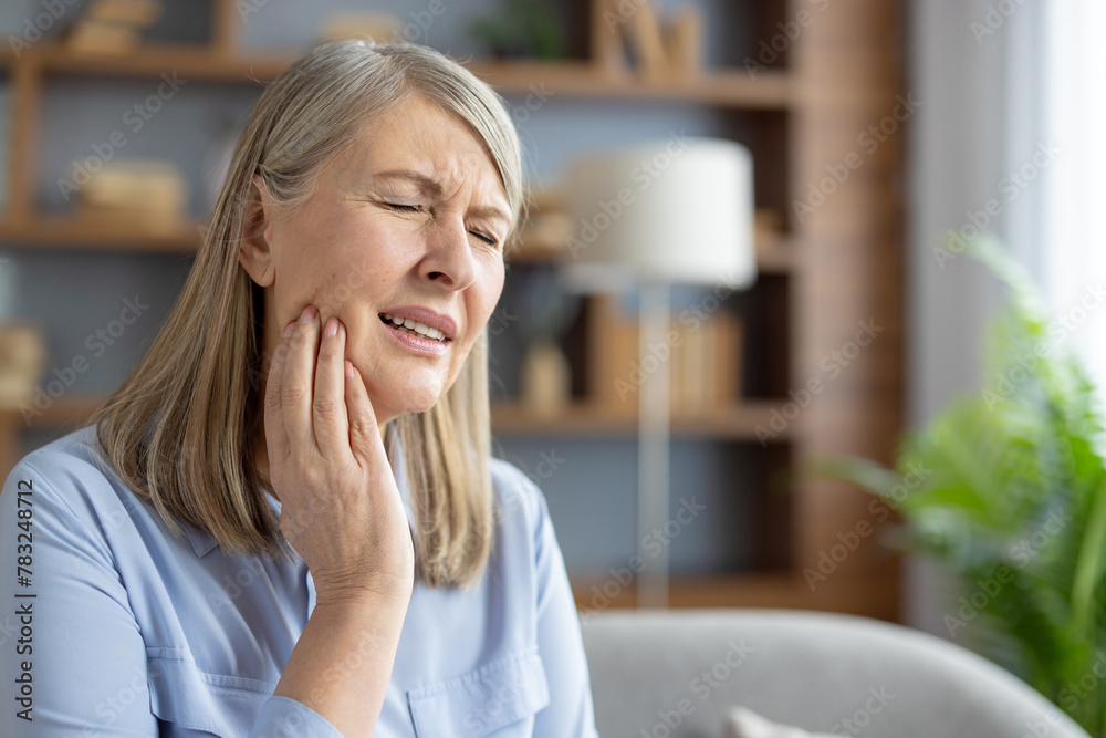 An elderly Caucasian woman sits at home, touching her cheek and ...