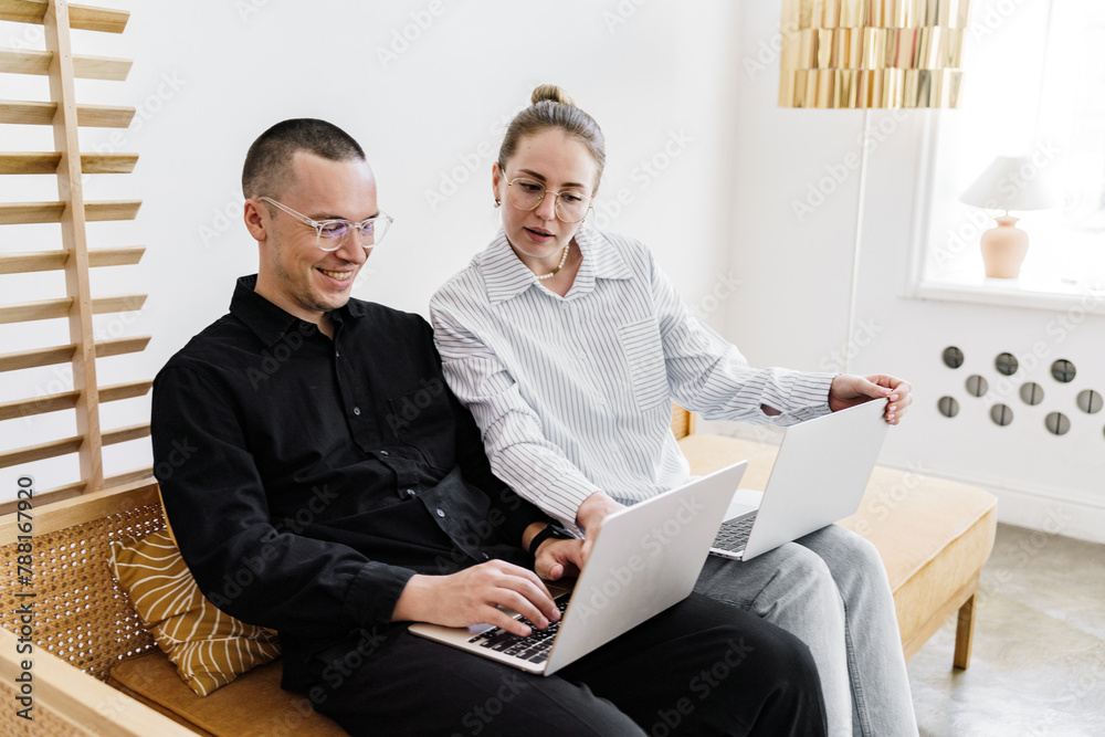 Two colleagues, one man and one woman, sit side by side, laptops in ...