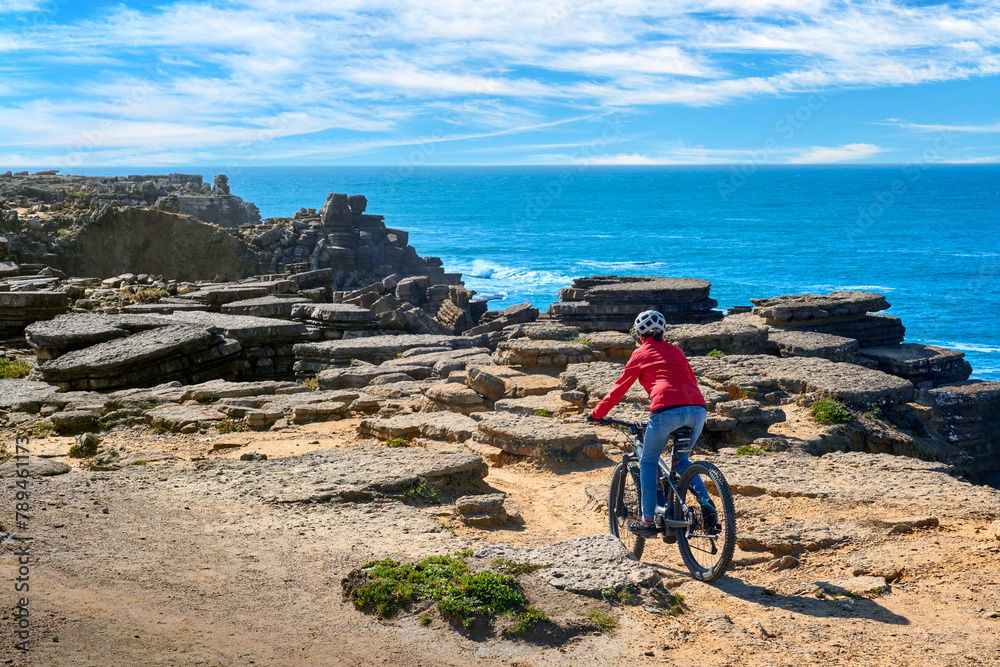 Brave senior woman riding her electric mountain bike on the rocky cliffs of Peniche at the ...
