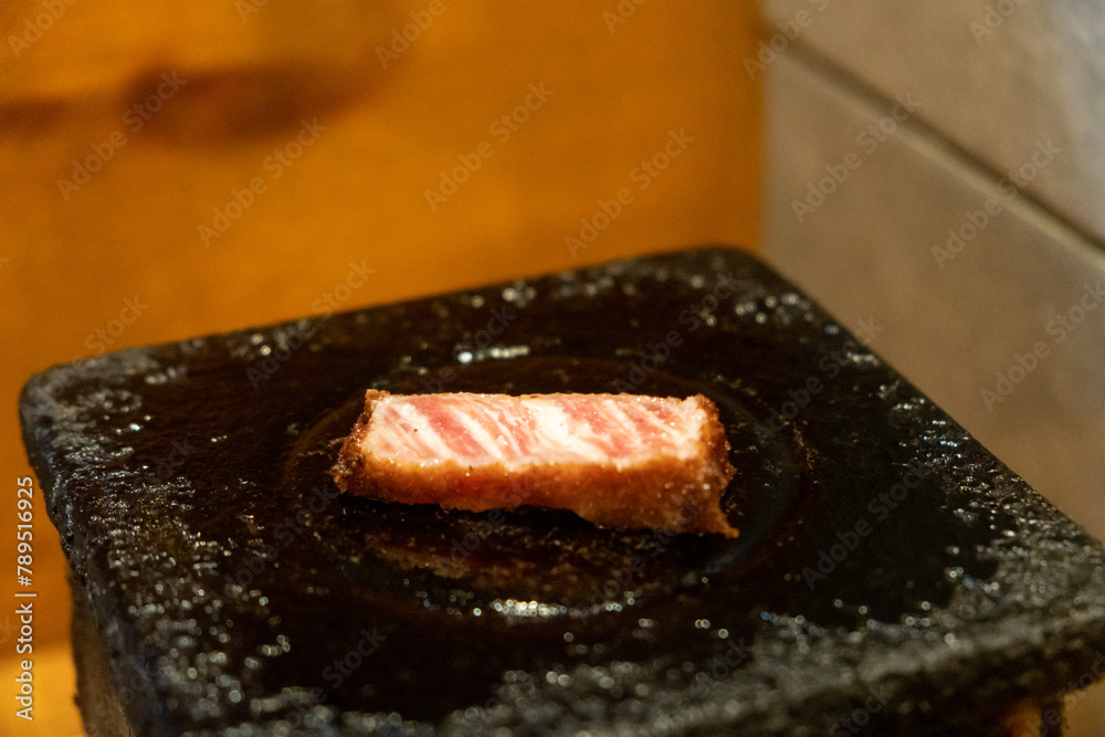 A piece of Gyukatsu or beef cutlet on the stone grill. It is ...