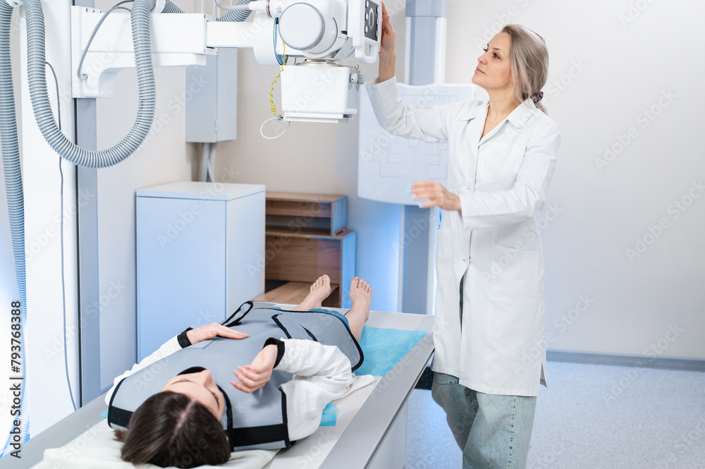 Female patient lying on bed while female nurse adjusting modern X-ray machine for scanning her ...